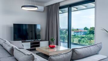 Uma sala de estar moderna com uma televisão de tela grande, sofá cinza e uma mesa de centro de vidro com um objeto decorativo vermelho.
