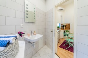 Um banheiro moderno com uma pia branca, um espelho e um chuveiro. É visível um porta-toalhas com toalha, e uma pequena planta está sobre o balcão.