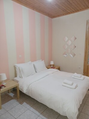 Cama bem arrumada com lençóis brancos, mesa de cabeceira com abajur e espelho fixado na parede.