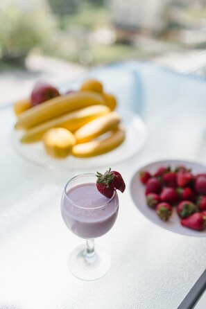 Uma mesa com um copo de bebida roxa decorado com morango, um prato de bananas e um prato de morangos.