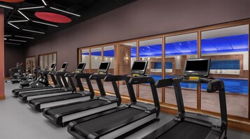 Centro de fitness com esteiras, bicicletas ergométricas e área de piscina visíveis por grandes janelas.