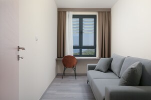 Uma sala de estar moderna com um sofá cinzento, uma poltrona castanha, uma janela com cortinas e uma porta.