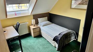 Quarto pequeno e compacto com cama, escrivaninha, cadeira, radiador e janela com vista para o exterior.