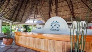 Uma área de recepção com teto de palha, um balcão de madeira e uma placa com a inscrição "Garoda Resort".