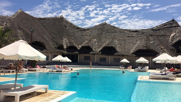 Piscina com espreguiçadeiras e guarda-sóis, cercada por edifícios com telhados de palha.