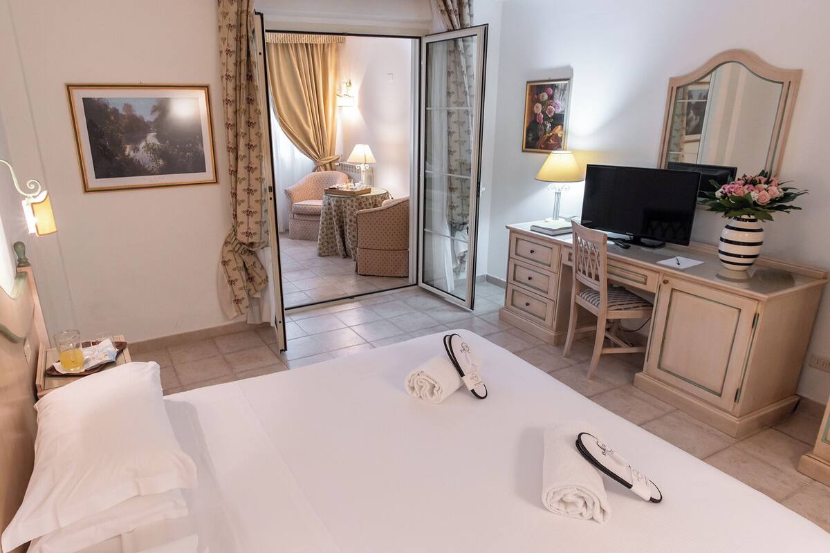 Quarto de hotel com cama, escrivaninha com televisão, cadeira, cômoda e porta para uma varanda.