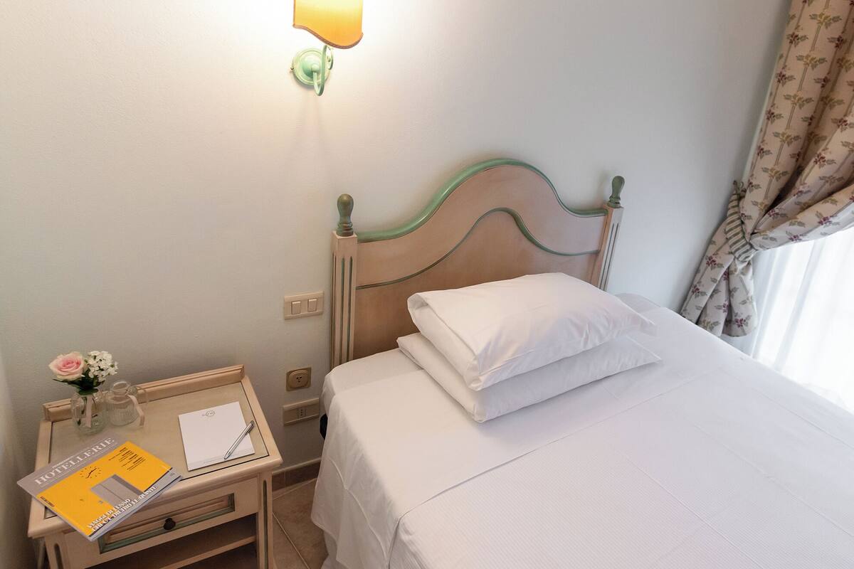 Quarto de hotel com cama, mesa de cabeceira com abajur, um vaso com flores e uma revista.