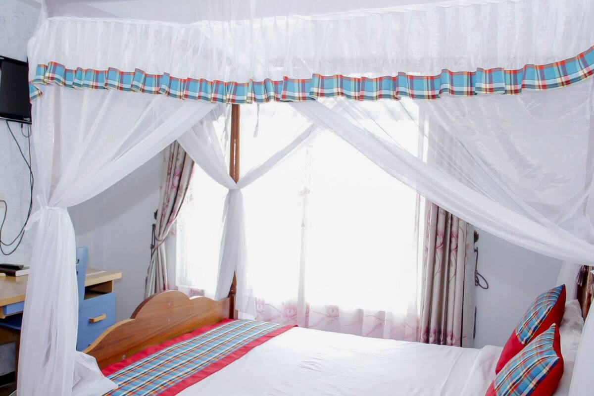 Quarto com cama sob um dossel branco, uma mesa de trabalho com cadeira e janela com cortinas.