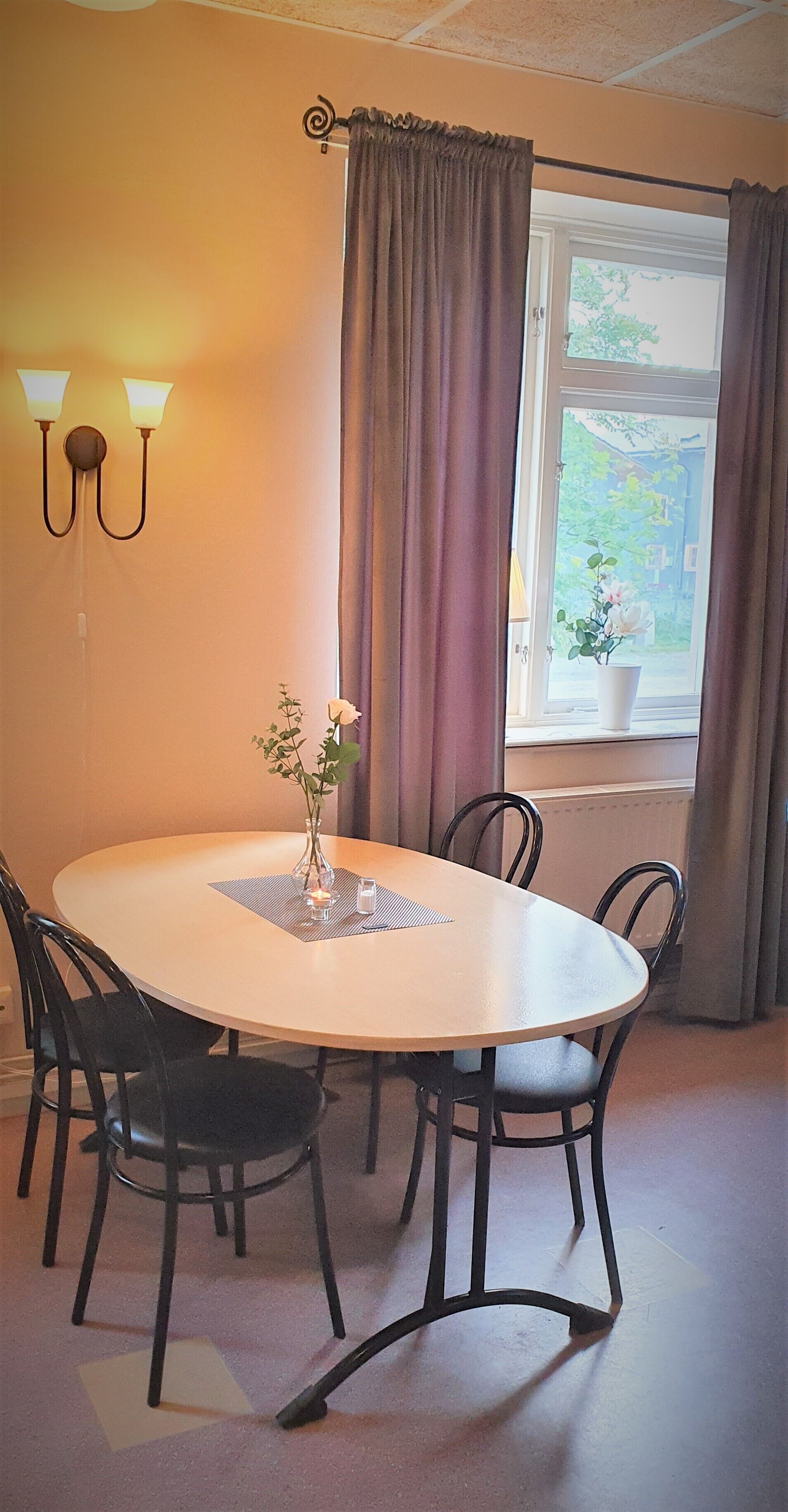Uma área de refeições com uma mesa redonda, cadeiras pretas, um vaso com flores e uma janela com cortinas.