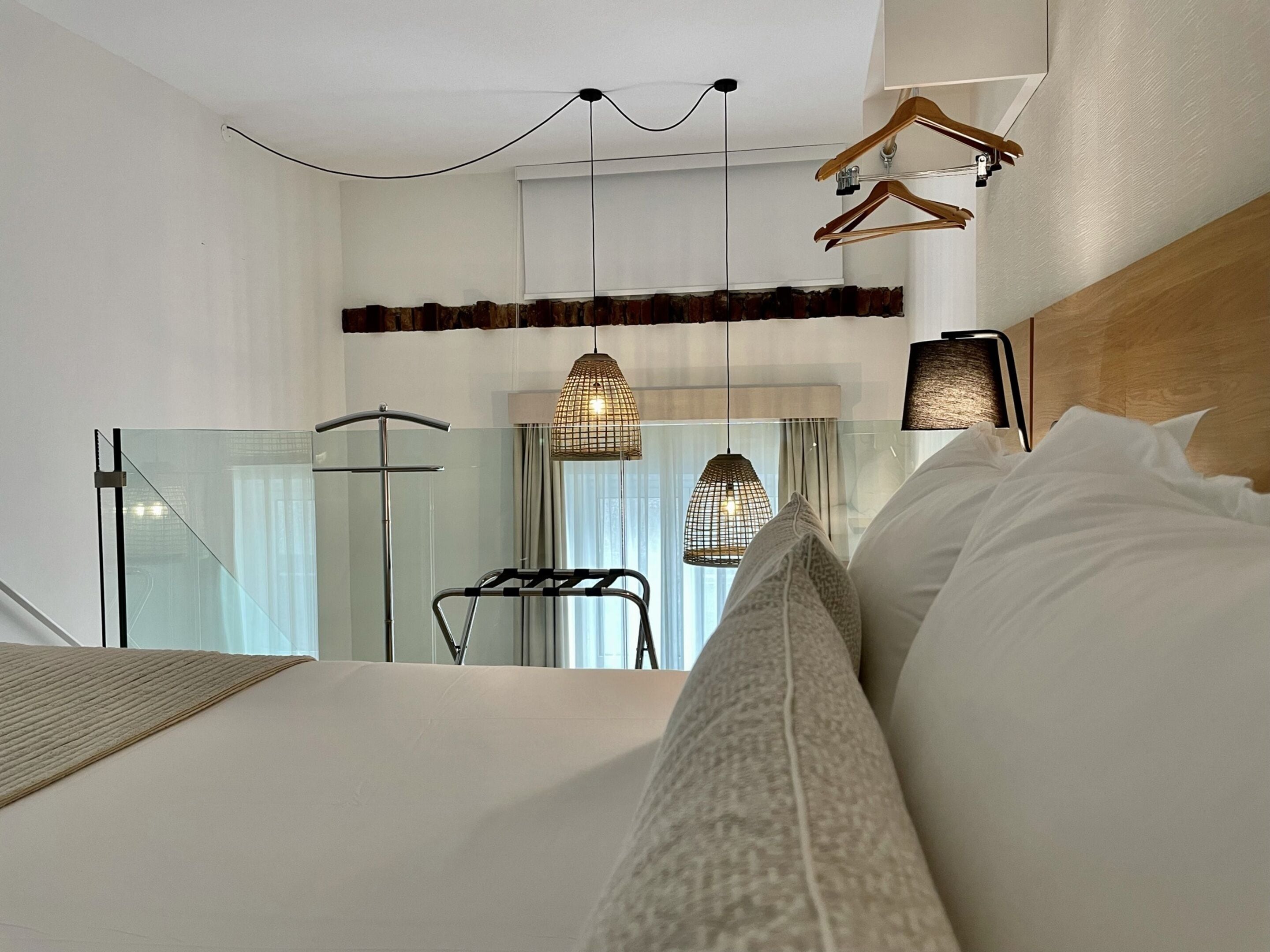 Quarto com cama, luminária pendente, cabeceira de madeira e vista para uma varanda com guarda-corpo de vidro.