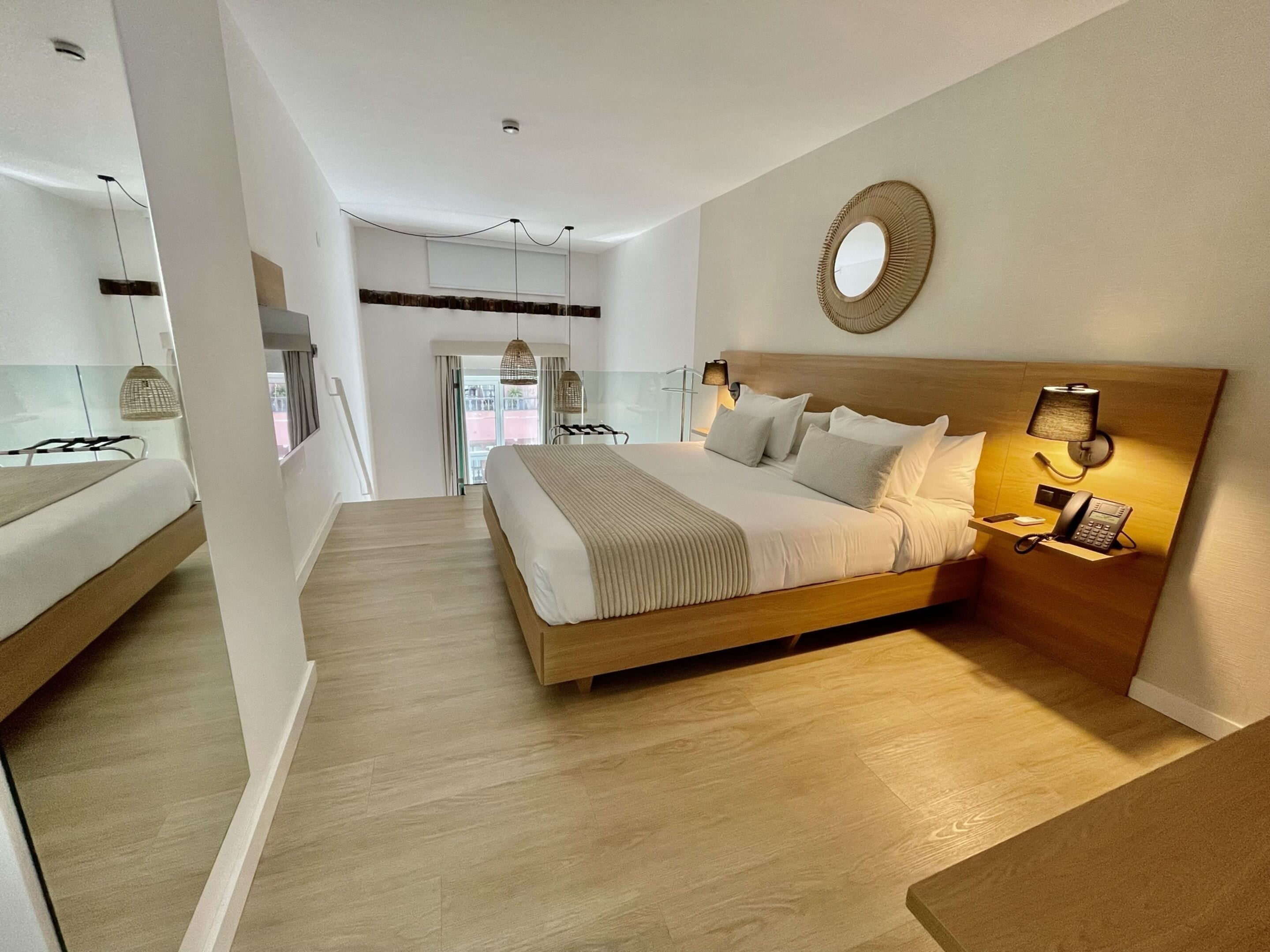 Quarto de hotel moderno com uma cama grande, luminárias laterais, um espelho e um telefone na mesa de cabeceira.