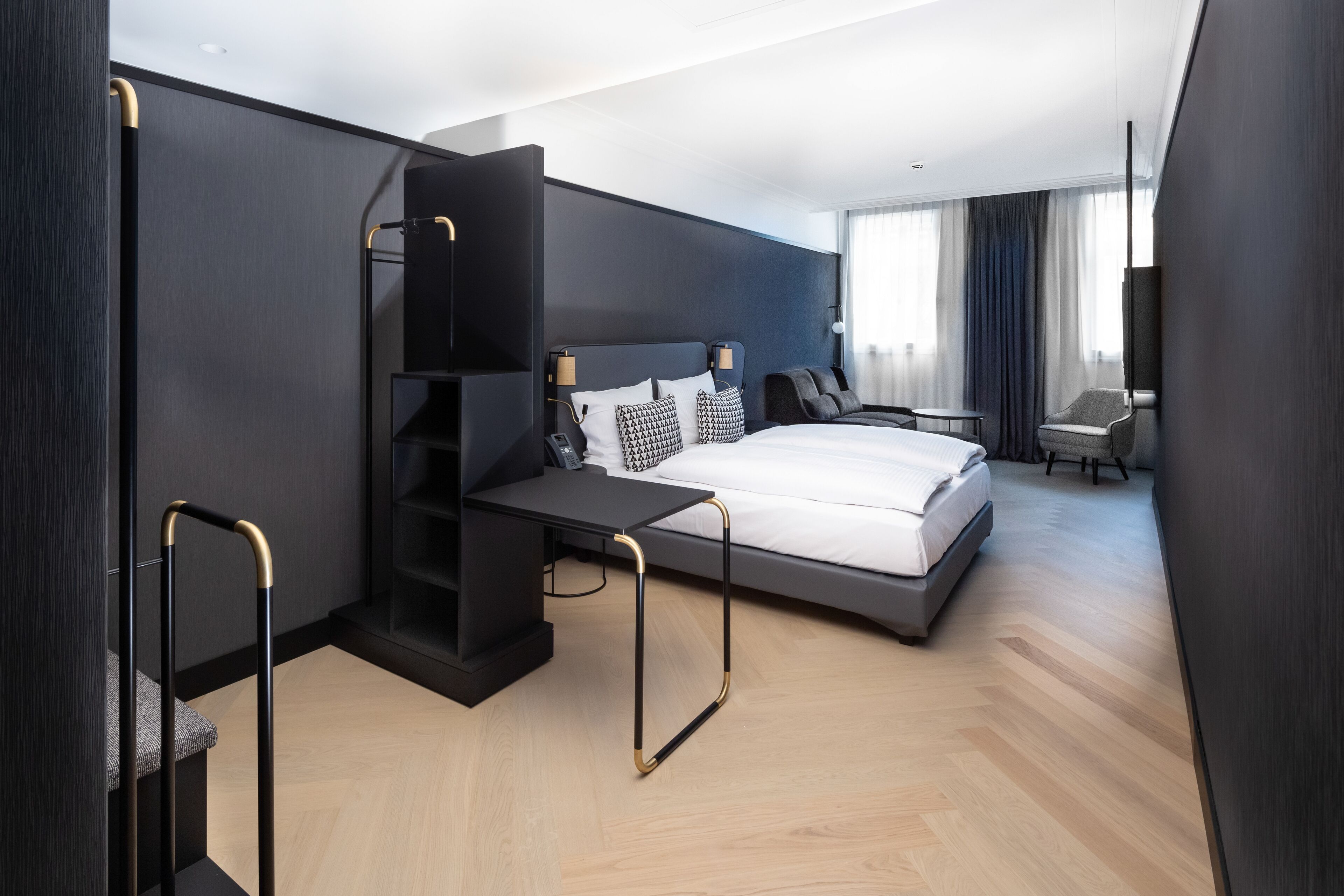 Quarto de hotel moderno com uma cama grande, uma escrivaninha com cadeira, uma mesinha e um sofá.