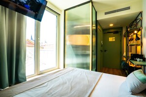 Quarto de hotel com cama, escrivaninha com luminária, uma cadeira e televisão fixada na parede.