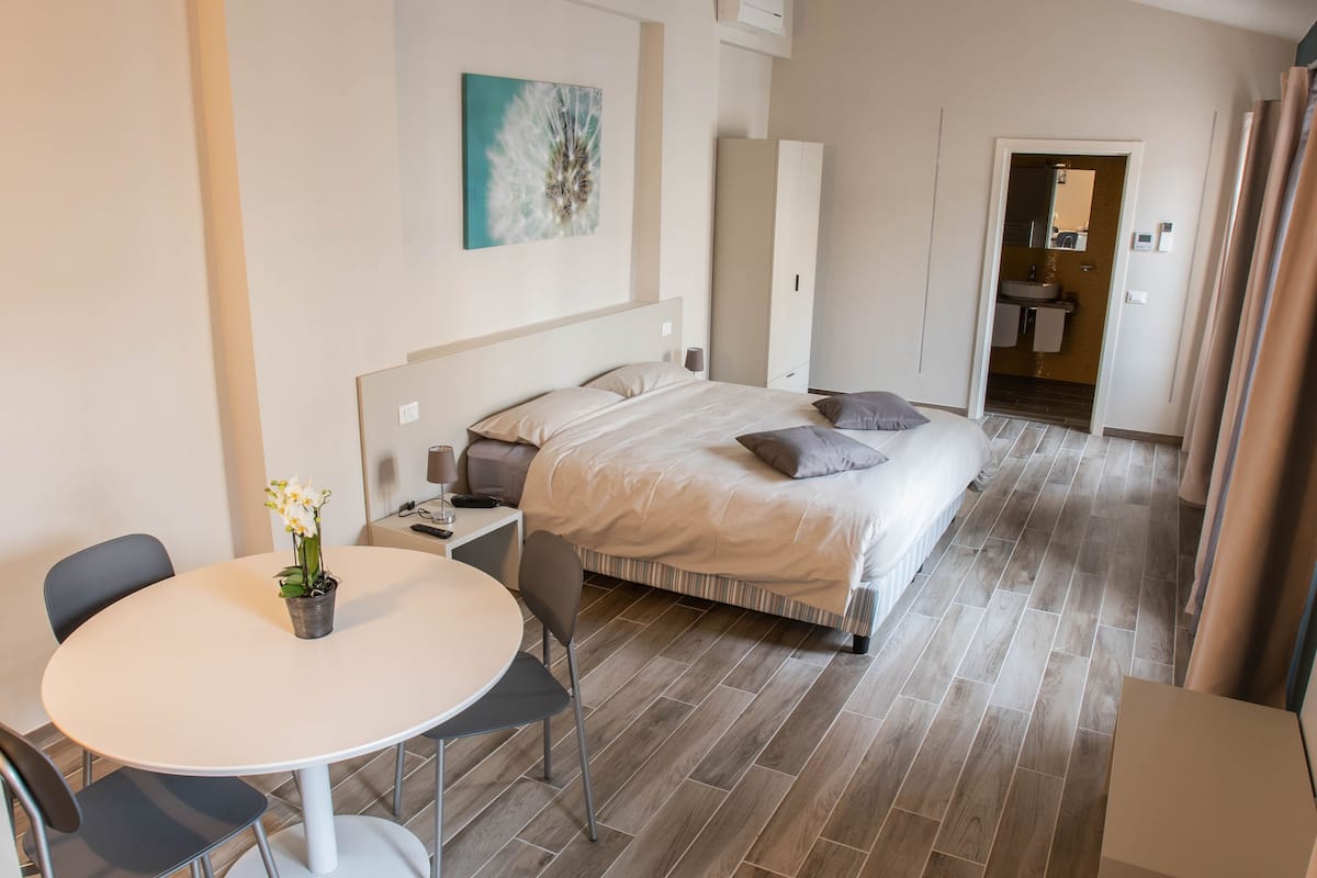 Quarto de hotel moderno com cama, mesa de cabeceira, abajur e uma pequena mesa com um vaso de flores.