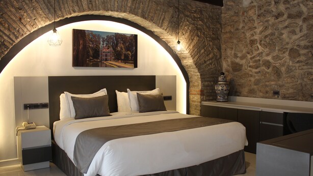 Quarto moderno com arco de pedra, cama com roupa de cama branca e cinza, mesa de cabeceira com um telefone e um quadro sobre a cama.
