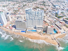 Paisagem costeira urbana com um hotel de vários andares em destaque, uma praia com guarda-sóis e uma vista clara do oceano.
