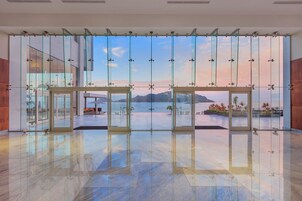 Um lobby amplo com uma grande porta de vidro que dá para um terraço ao ar livre, com vista para montanhas e um corpo d'água.