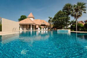 Piscina azul cristalina com um deck ladrilhado, rodeada por palmeiras e um edifício com telhado em azulejos.