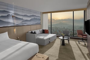 Quarto de hotel com cama, sofá, mesa pequena, cadeira e uma janela grande com vista para a cidade.