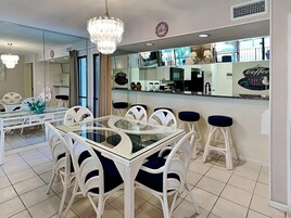 Uma área de refeições com uma mesa de vidro rodeada por cadeiras brancas com almofadas azuis, um candelabro acima e uma cozinha visível através de um painel de vidro.