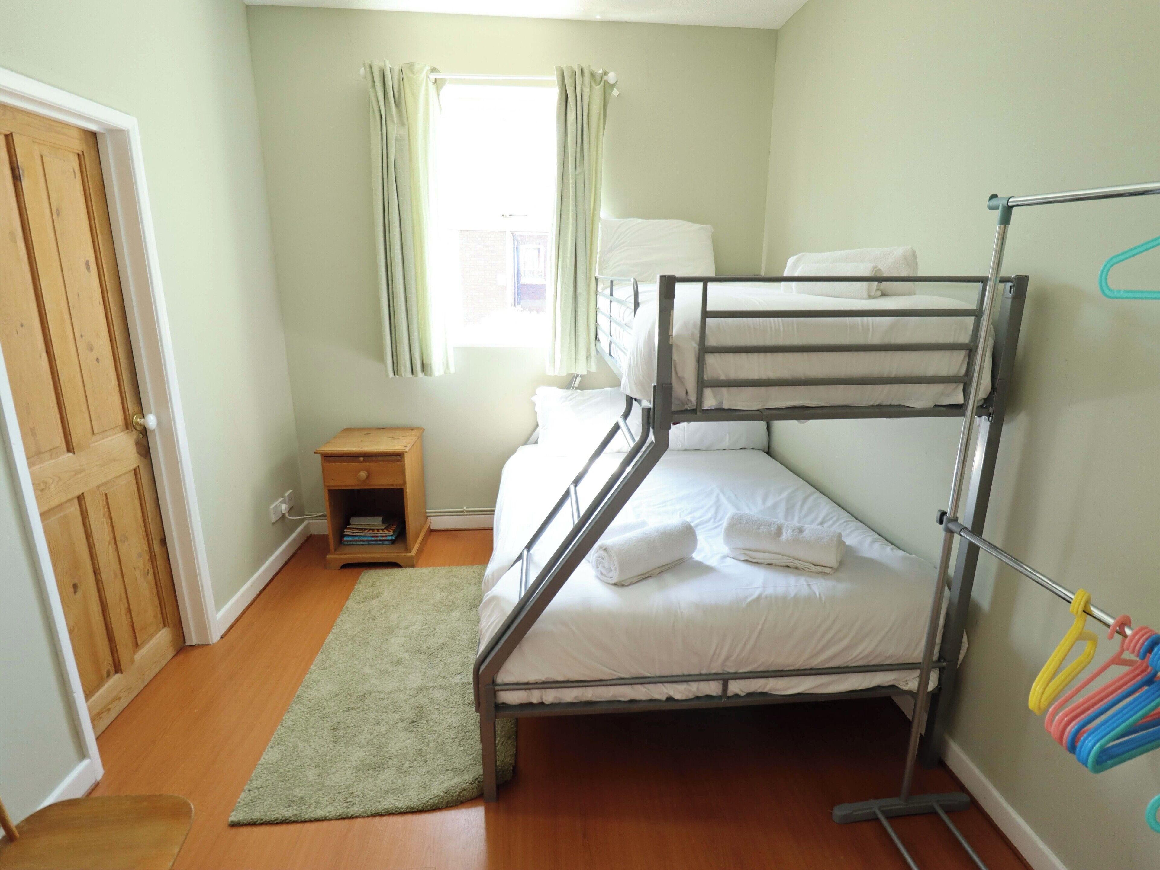 Quarto com beliche, porta de madeira, mesinha de cabeceira, tapete verde e janela com cortinas.