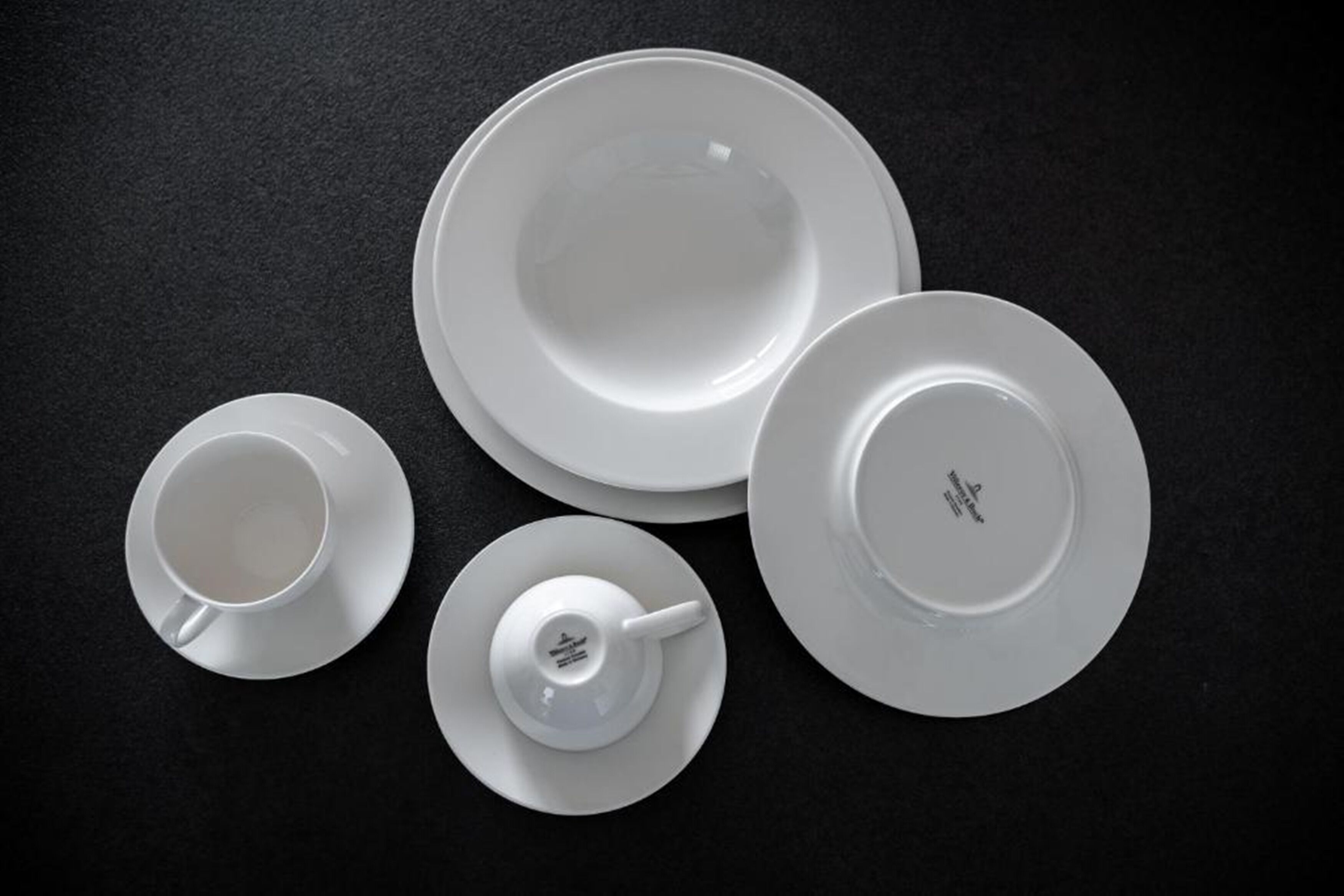Um conjunto de pratos de cerâmica branca, uma chávena e um pires dispostos sobre uma superfície escura.