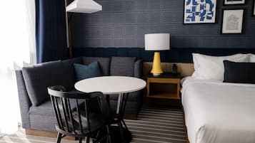 Quarto de hotel com uma cama, um sofá, uma pequena mesa redonda, uma cadeira, um abajur e uma parede com obras de arte.