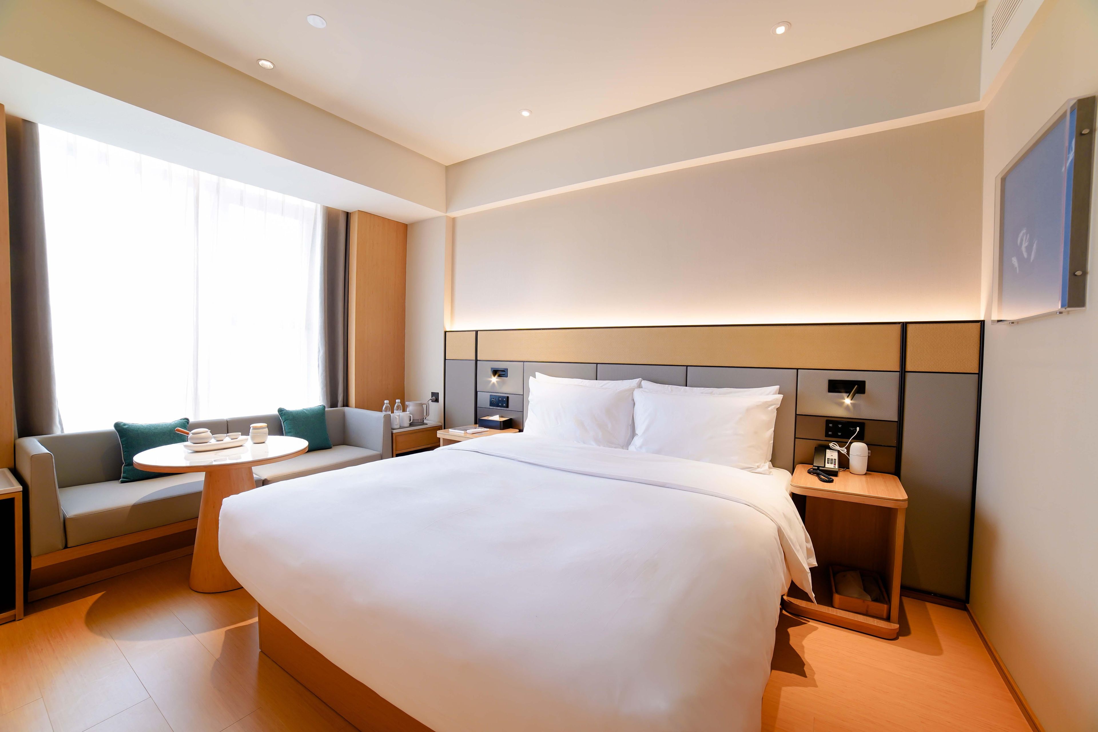 Quarto de hotel com uma cama grande, mesa de cabeceira, mesa redonda pequena com uma cadeira, sofá e uma janela ampla.