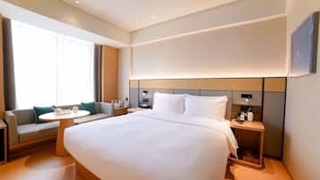 Quarto de hotel com uma cama grande, mesa de cabeceira, mesa redonda pequena com uma cadeira, sofá e uma janela ampla.