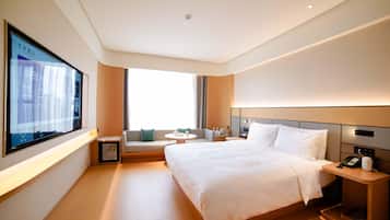 Quarto de hotel moderno com uma cama grande, mesa de cabeceira com telefone e abajur, um pequeno espaço de estar com sofá e mesa, e uma grande janela que oferece luz natural.