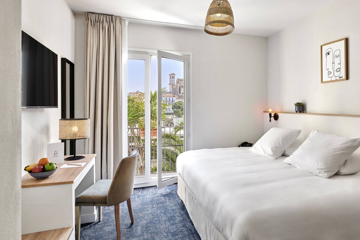 Quarto de hotel com cama, escrivaninha, cadeira e vista para a cidade pela janela.
