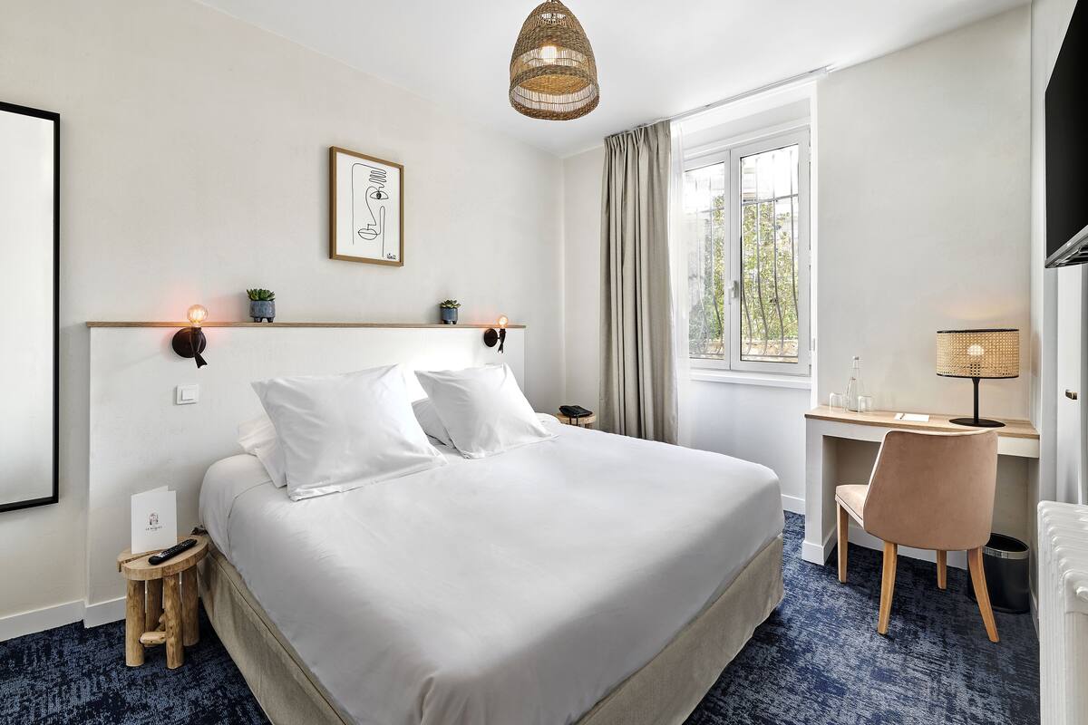 Quarto de hotel com uma cama grande, uma escrivaninha, uma cadeira, um candeeiro pendente e uma janela com cortinas.