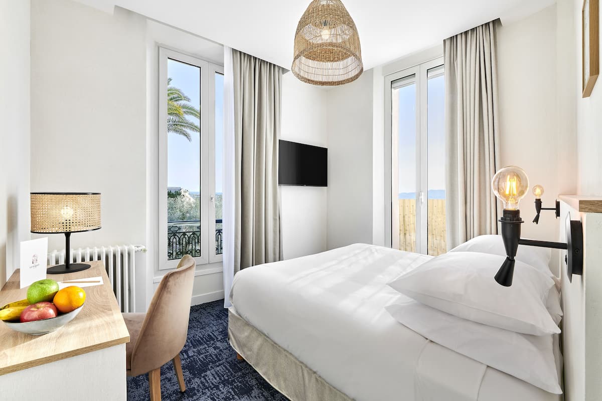 Quarto de hotel com cama, secretária com luminária, cesta de frutas e vista para o exterior.