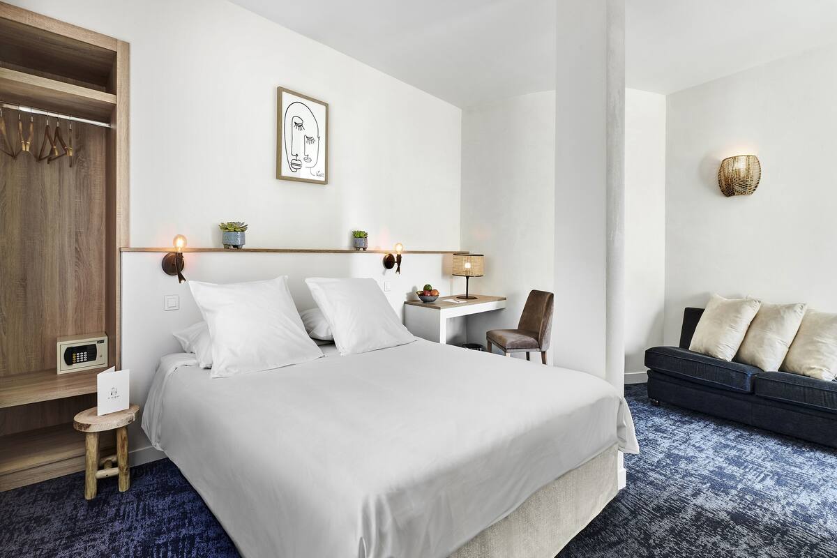 Quarto de hotel com cama, escrivaninha, poltrona, sofá, guarda-roupa e luminária fixada na parede.