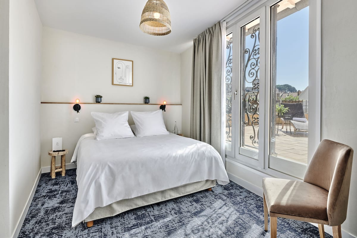 Quarto de hotel com cama, uma cadeira, janela com cortinas, luminária na parede e iluminação no teto.