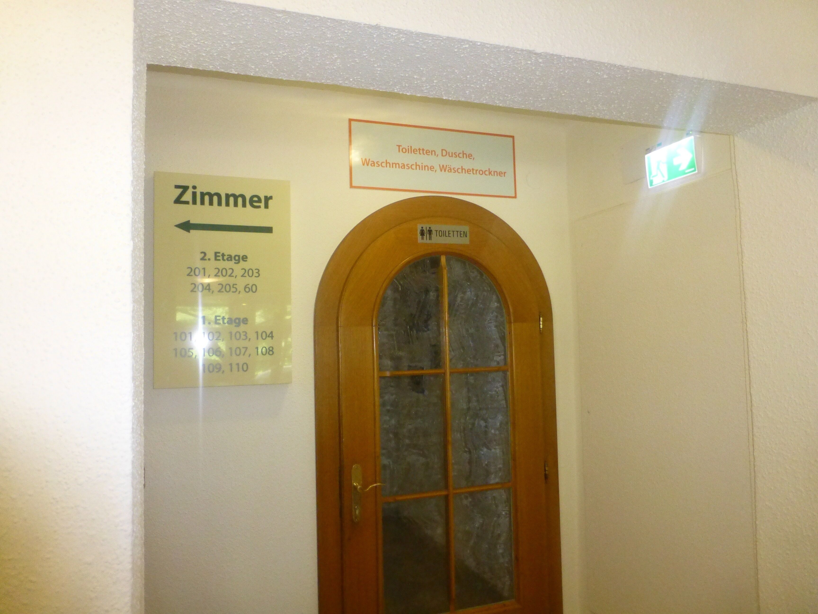 Entrada de um quarto de hotel com porta de madeira, placa indicando o tipo de quarto e camas disponíveis, e uma seta apontando para a esquerda.