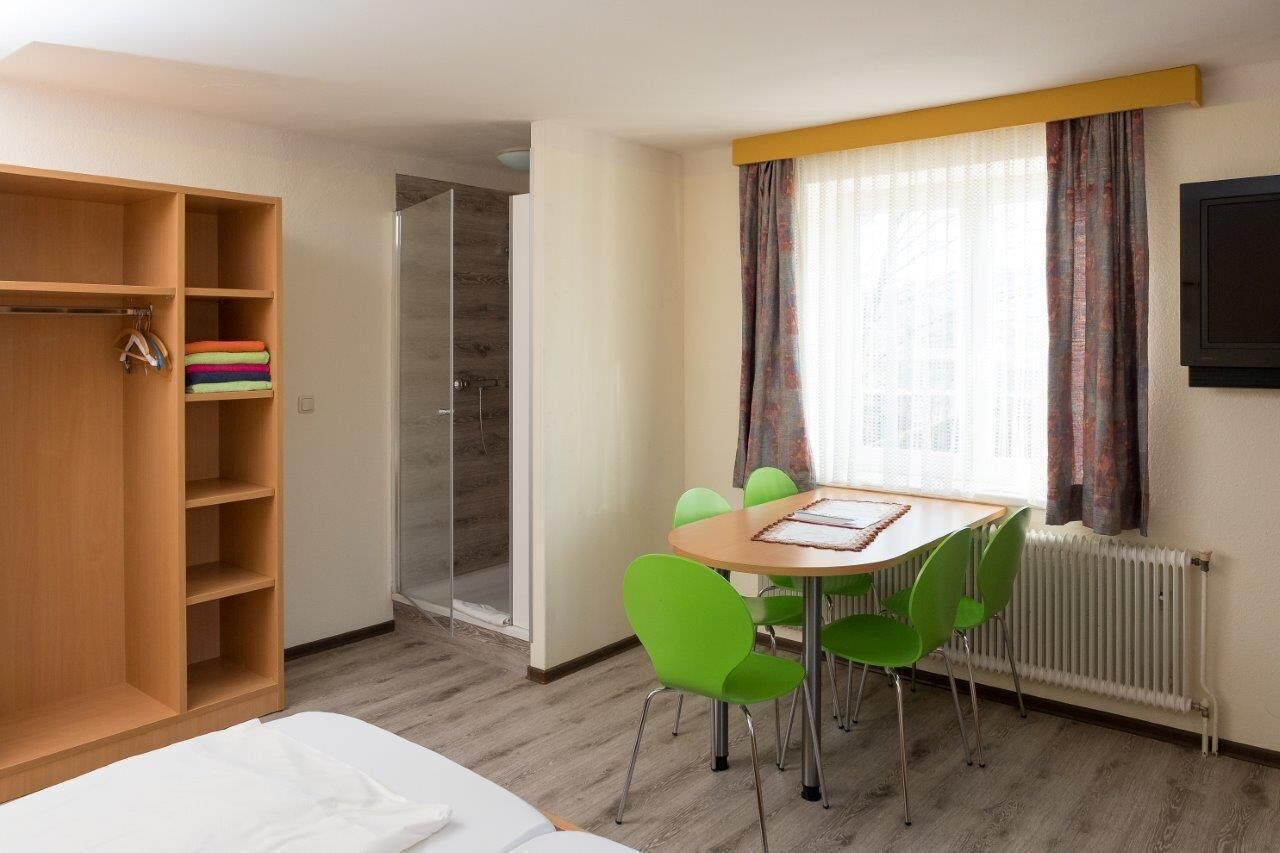 Quarto de hotel com guarda-roupa de madeira, uma cama, uma mesa redonda com cadeiras verdes, chuveiro e janela com cortinas.