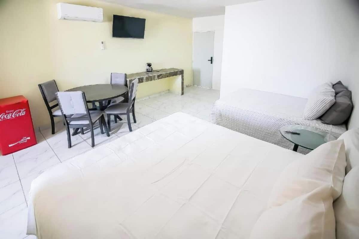 Quarto de hotel com uma cama, uma mesa de jantar com cadeiras, uma mesinha, um sofá, uma TV fixada na parede e um cooler vermelho Coca-Cola.