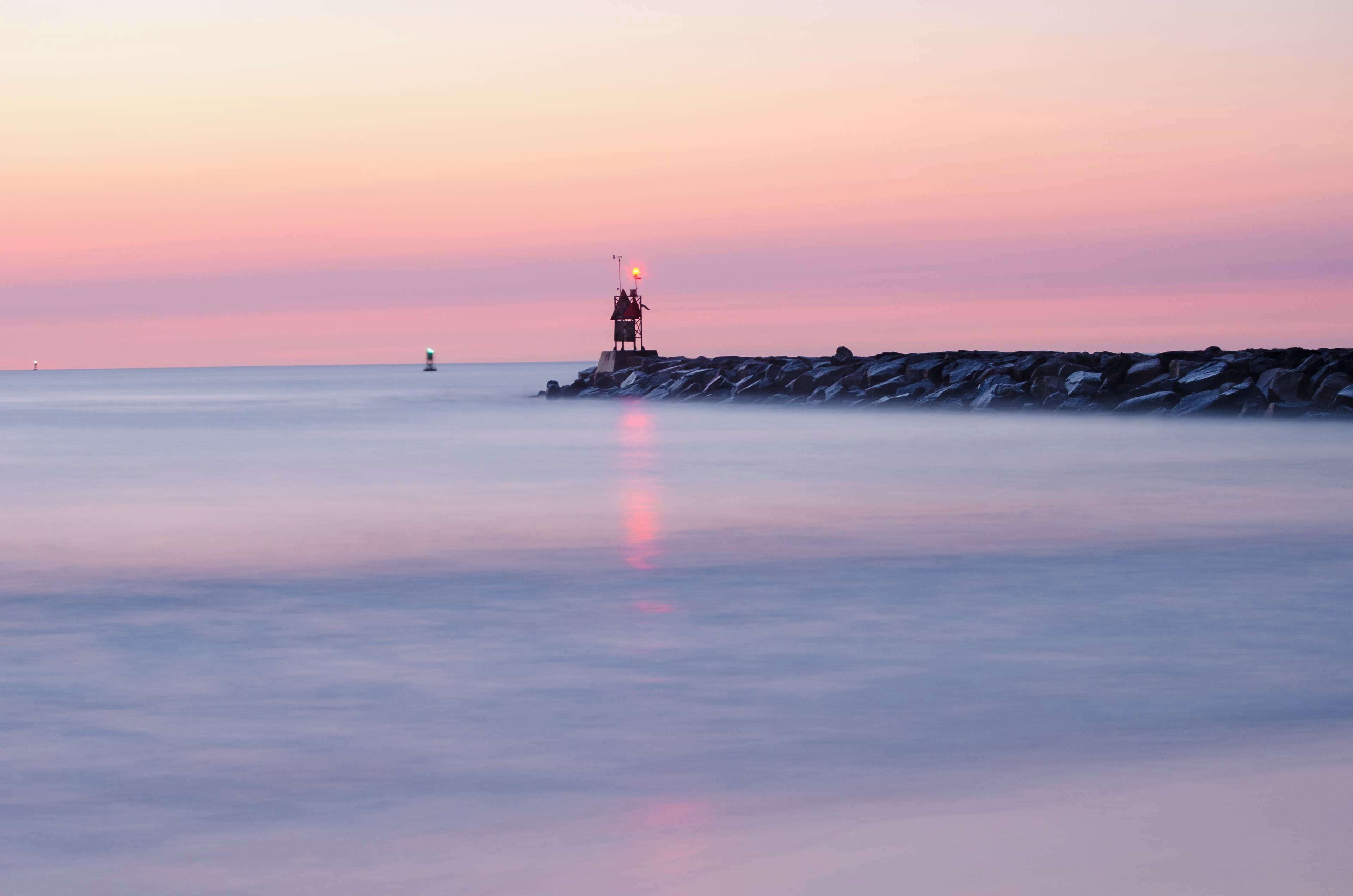 Mar calma ao pôr do sol com um farol numa formação rochosa.