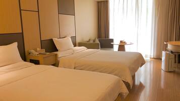 Quarto de hotel com duas camas, uma escrivaninha, uma cadeira, uma mesinha e uma janela grande com cortinas.