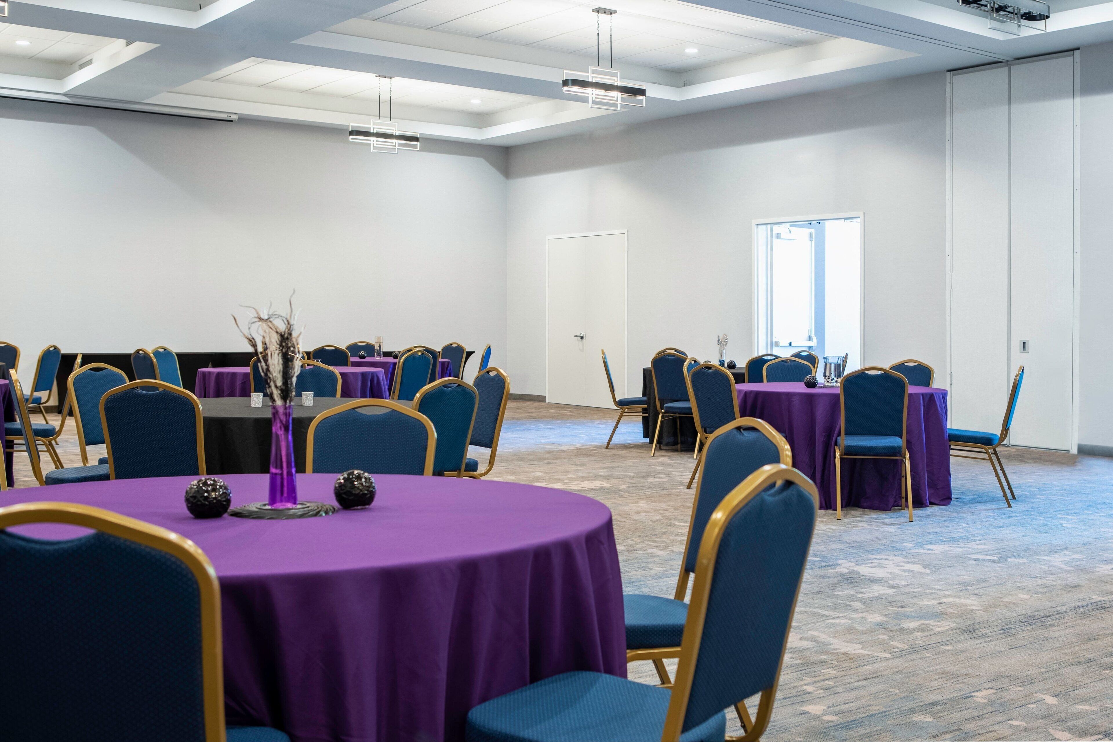 Sala de conferência com mesas redondas cobertas por toalhas roxas, cadeiras azuis e tela de projeção.