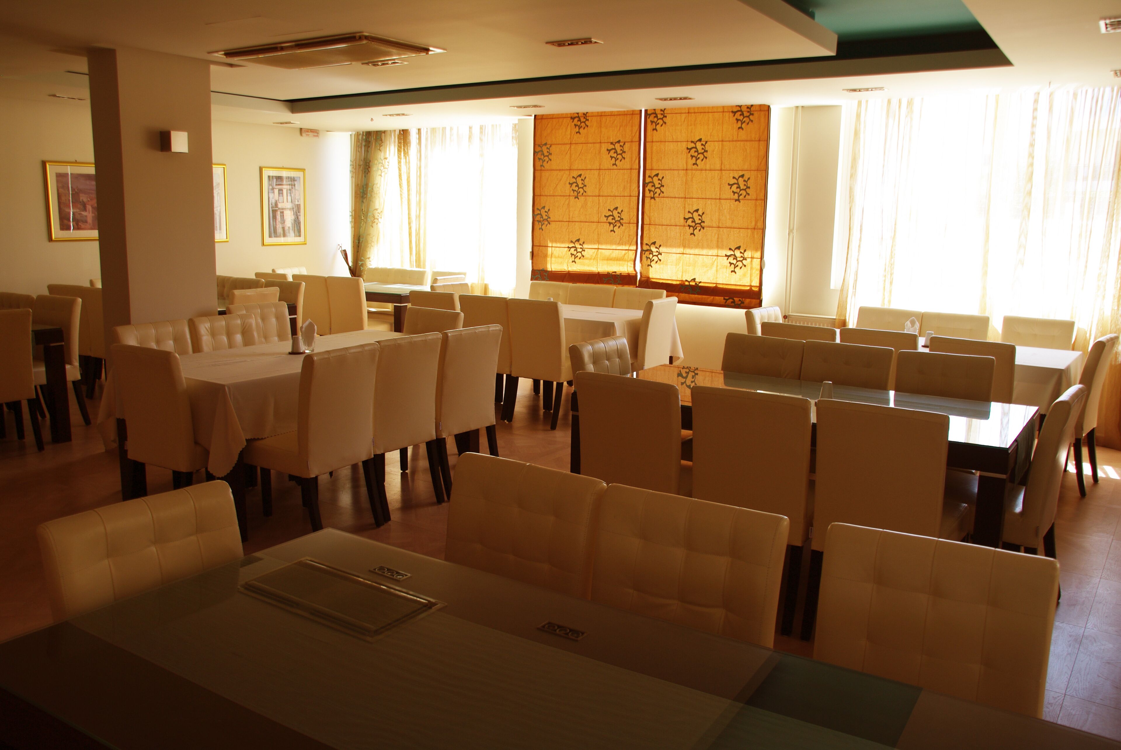 Uma área de refeições de hotel com várias mesas e cadeiras, grandes janelas com cortinas e quadros nas paredes.