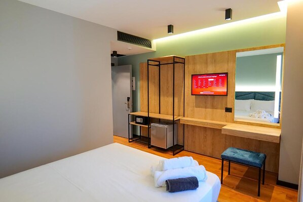 Quarto de hotel moderno com cama, pequena escrivaninha, espelho e televisão exibindo um cardápio.