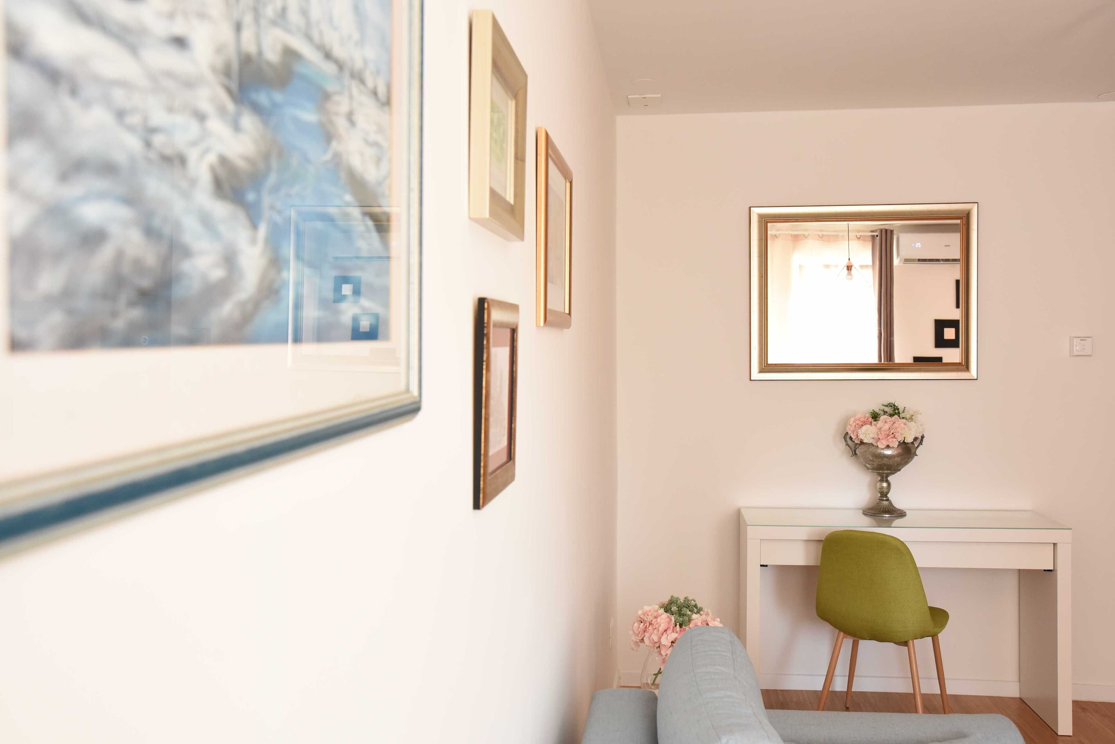 Quarto com escrivaninha branca, cadeira verde, quadro emoldurado e um vaso com flores.