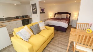 Quarto de hotel compacto com sofá amarelo, mesa de jantar de madeira, cama com colcha estampada, pequena cozinha e obra de arte pendurada na parede.