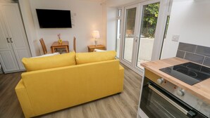 Cozinha compacta com sofá amarelo, mesa de jantar de madeira, TV fixada na parede e porta de vidro corrediça que dá para uma área externa.