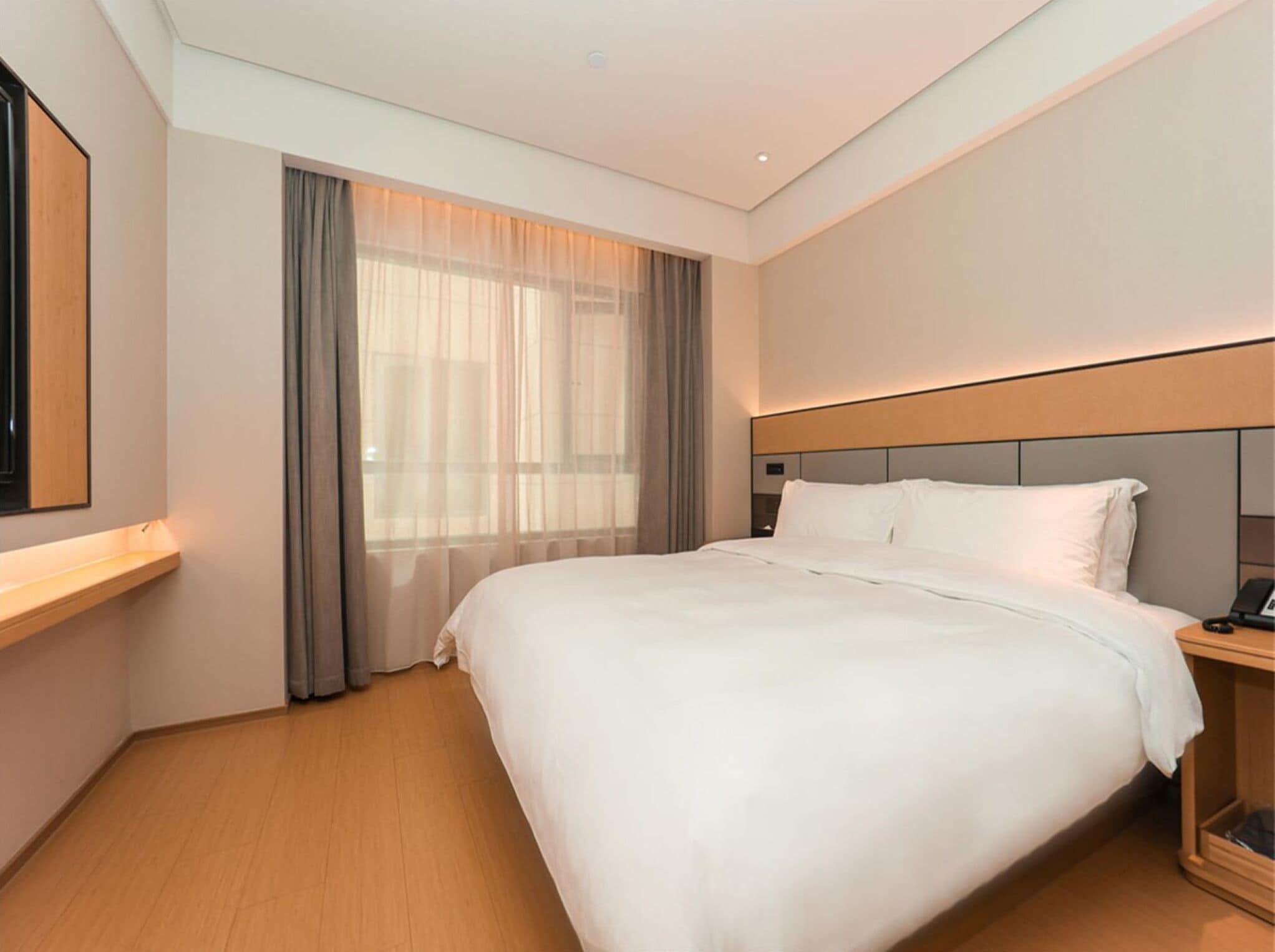 Quarto de hotel com uma cama grande, mesa de cabeceira com telefone, janela com cortinas e TV fixada na parede.