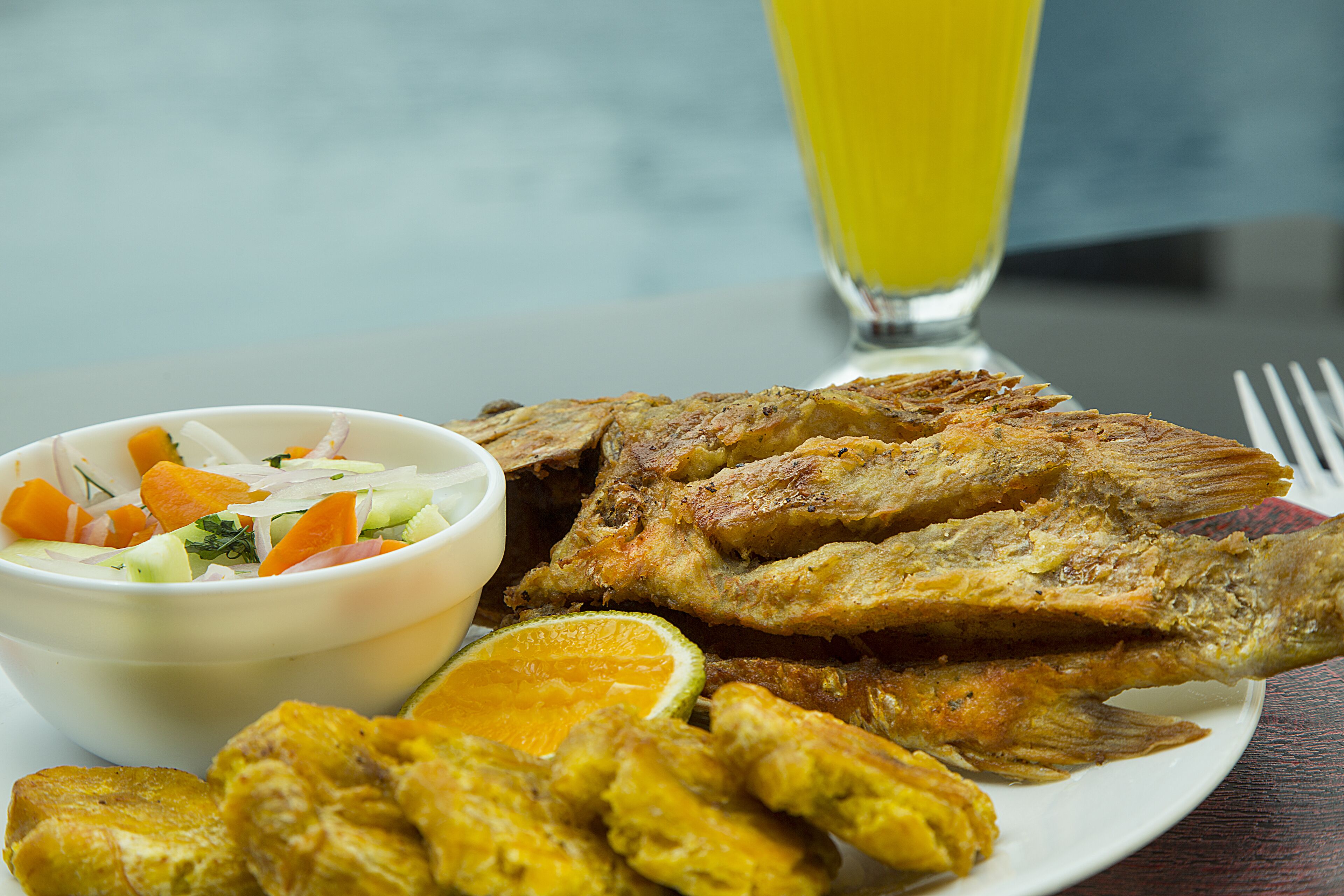 Um prato de peixe frito e banana da terra frita, acompanhado de uma salada e um copo de suco de laranja.