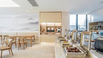 Uma área de refeições moderna em hotel, com buffet, mesas e vista para a cidade.