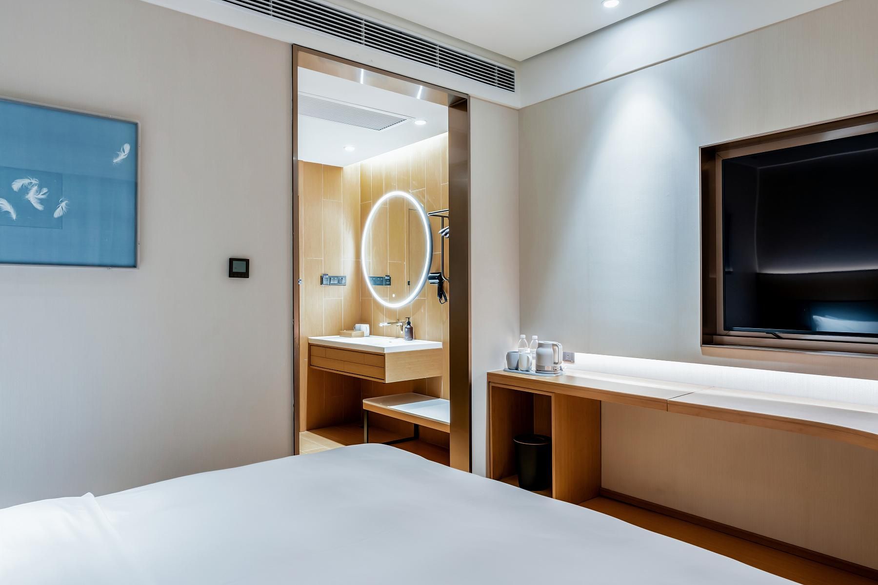 Quarto de hotel com cama, espelho grande iluminado, televisão e banheiro com pia e chuveiro.